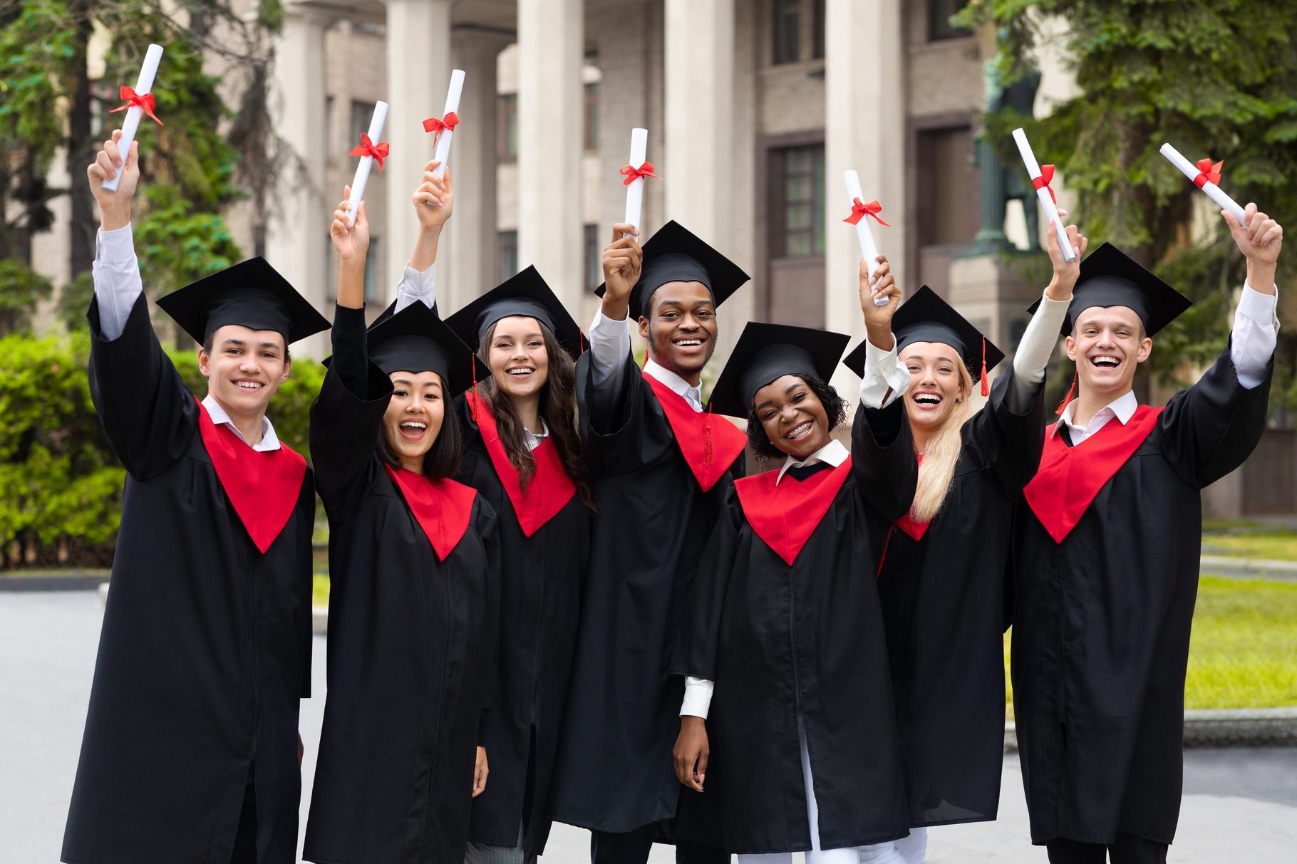 Cheerful multiracial students in graduation costumes raising diplomas ...